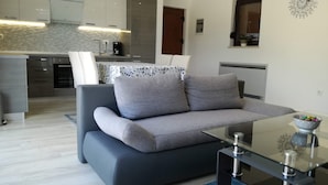 Uma sala de estar moderna com um sofá cinzento, uma mesa de centro de vidro e uma cozinha ao fundo.