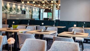Interior de restaurante com várias mesas e cadeiras, uma luminária pendente grande e um mural na parede.