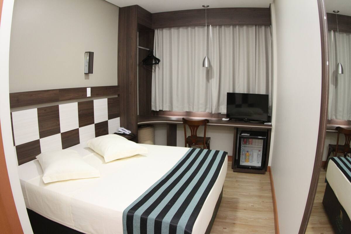Quarto de hotel com uma cama, escrivaninha, televisão e mini-geladeira.
