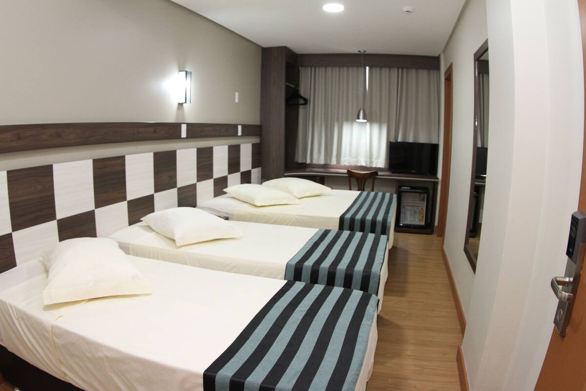 Quarto de hotel com três camas, uma televisão e uma escrivaninha com cadeira.