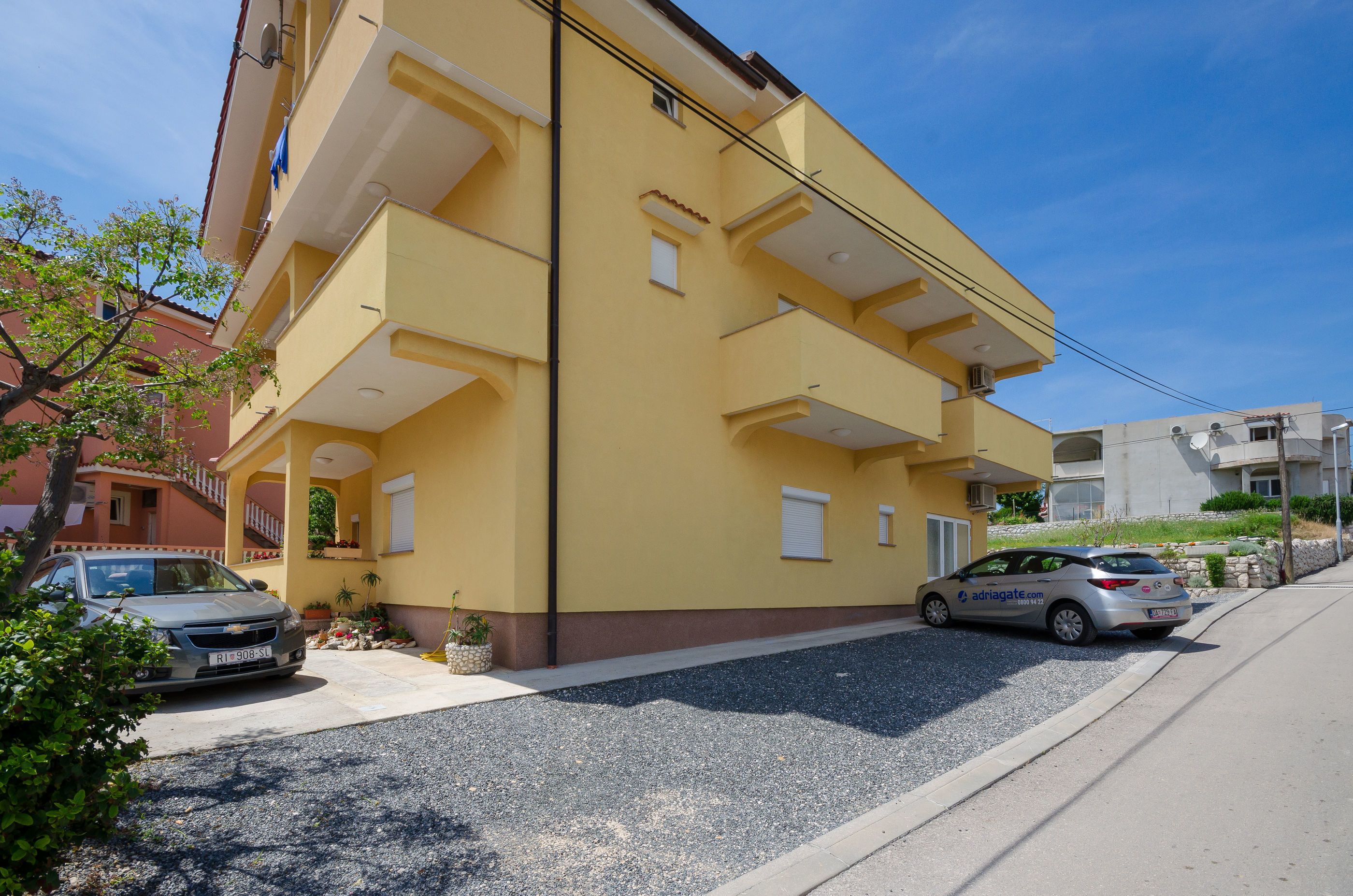 Um edifício residencial amarelo de vários andares com varandas, um carro estacionado em frente e um céu azul límpido.
