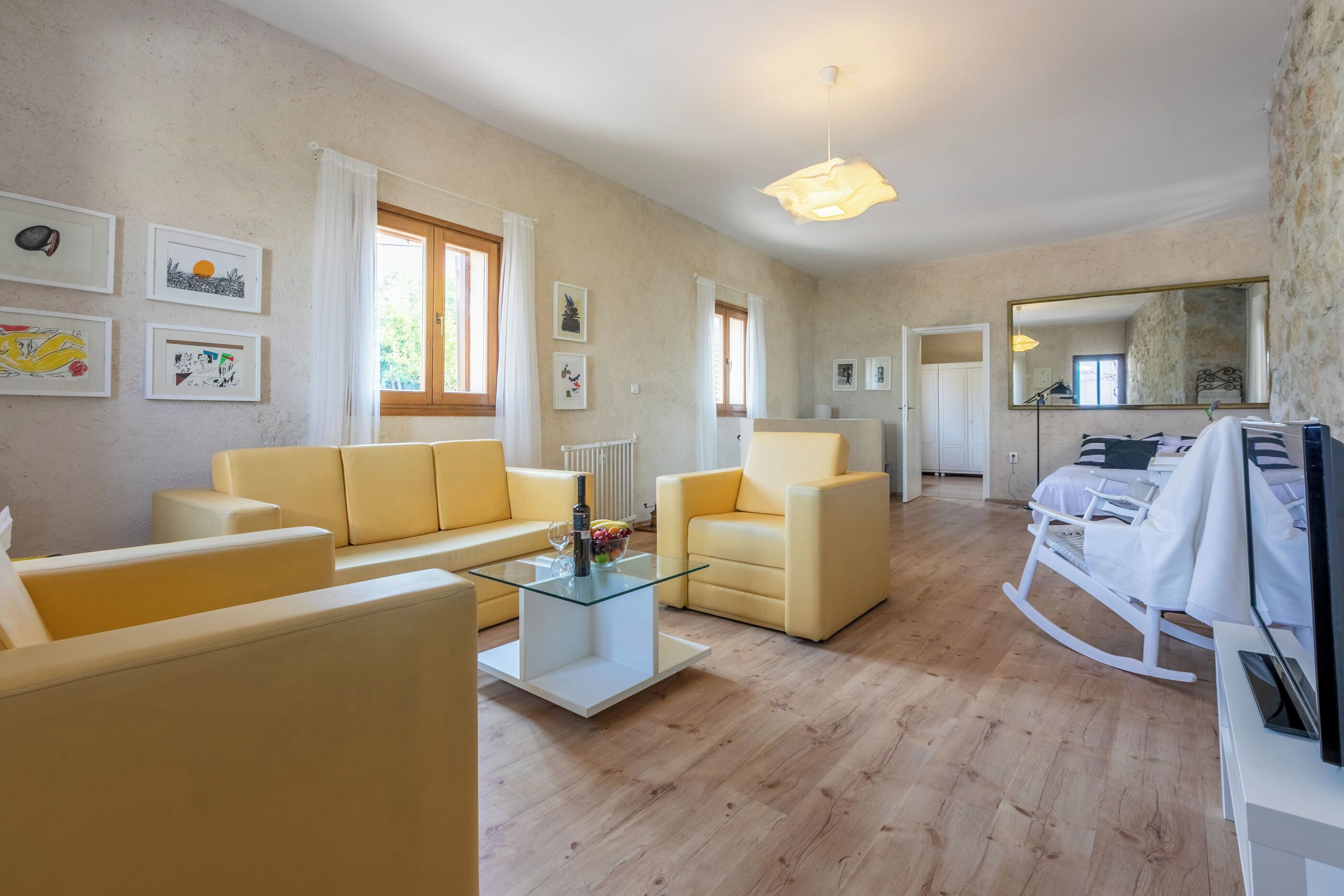 Uma sala de estar moderna com sofá, poltrona e mesa de centro de vidro. Há quadros nas paredes e um espelho grande.