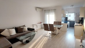 Uma sala de estar moderna com uma mesa de centro de vidro, um sofá com almofadas, uma área de jantar com cadeiras e uma cozinha equipada.