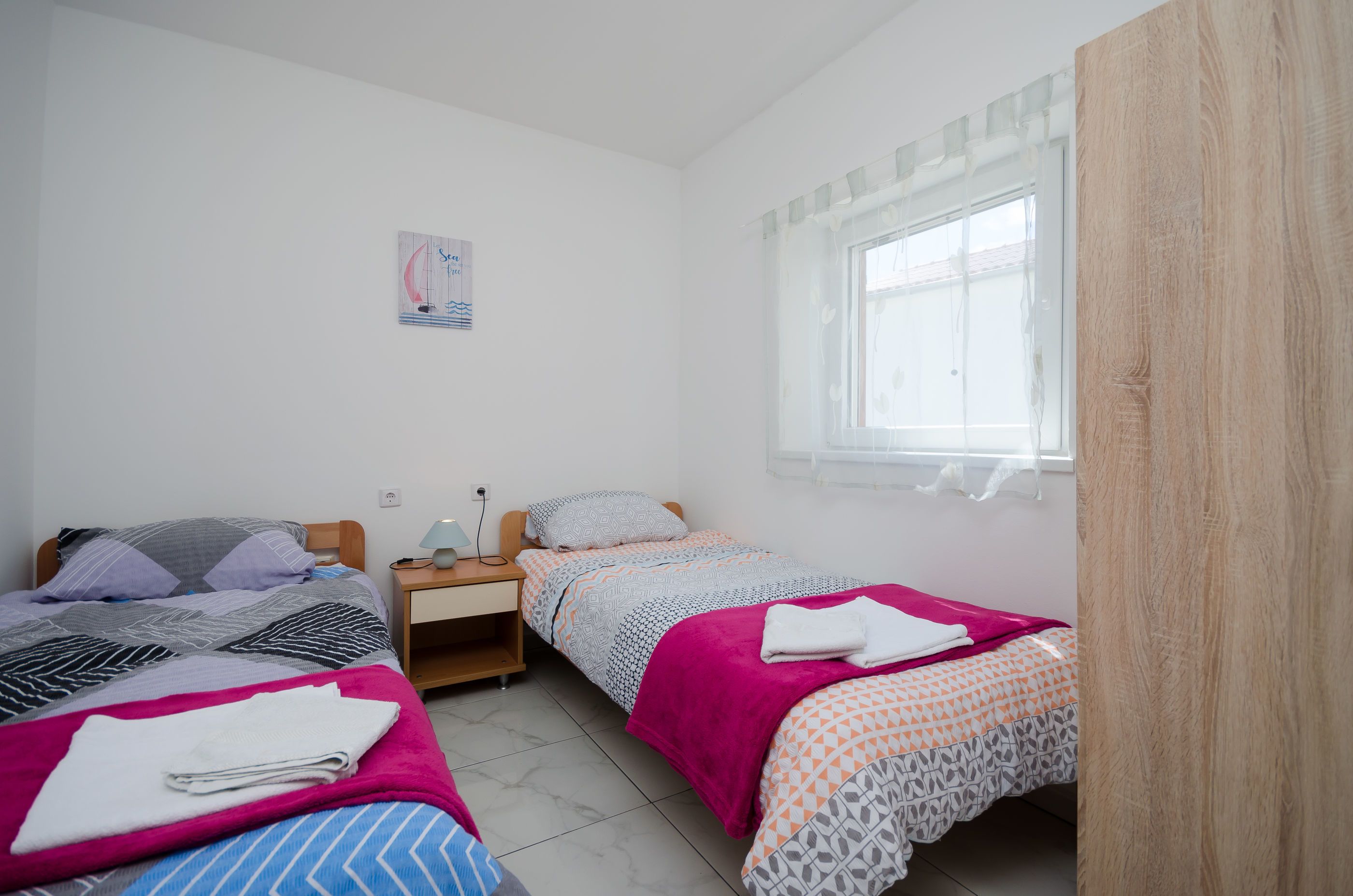 Quarto pequeno e limpo, com duas camas, uma mesa de cabeceira e uma janela com cortinas finas.