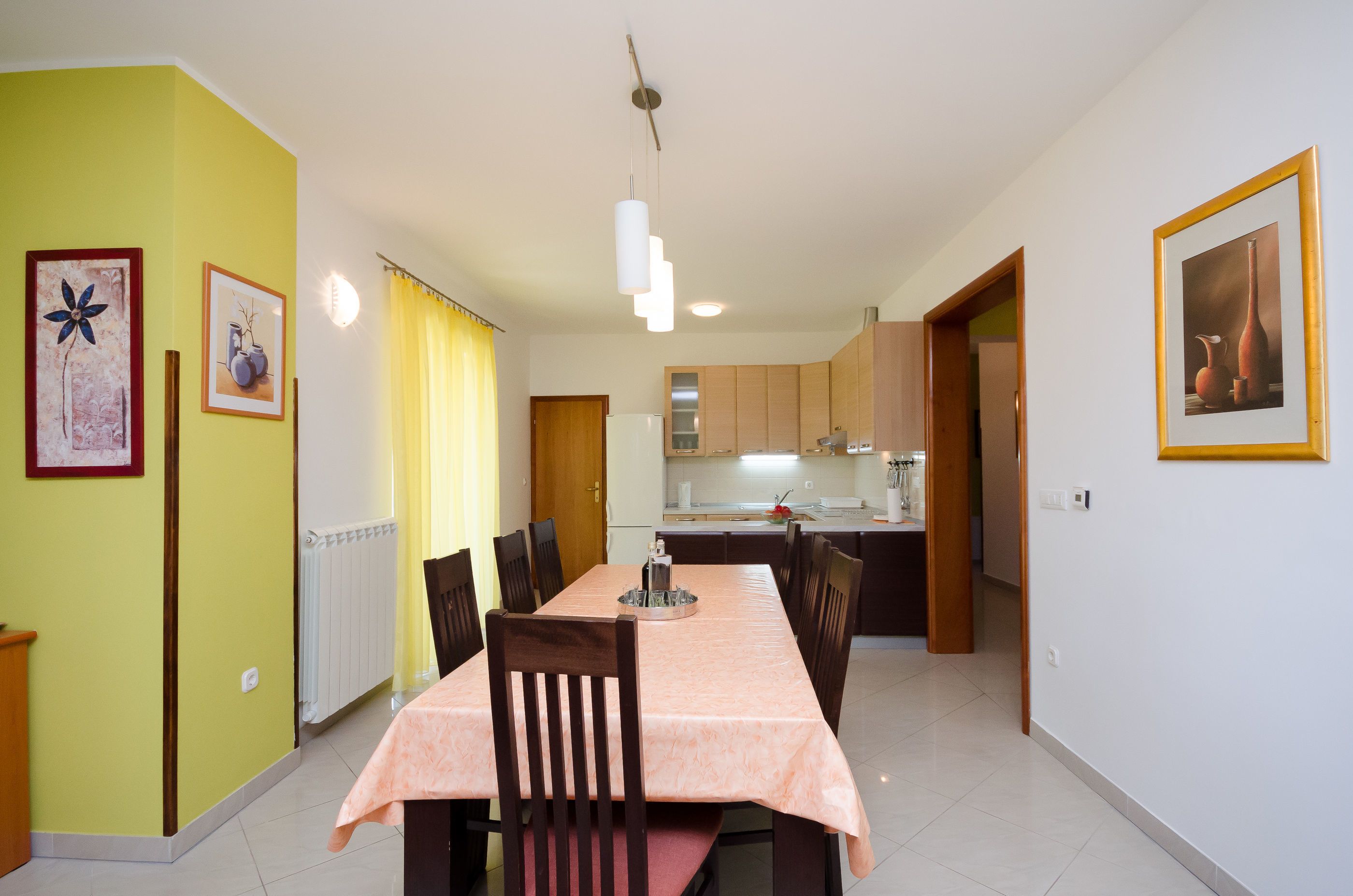 Uma sala de jantar com uma mesa posta para quatro pessoas, cadeiras de madeira, um lustre e armários de cozinha ao fundo.
