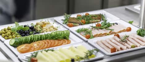 Buffet com diversos alimentos, incluindo pepino fatiado, tomate, queijo e carnes.