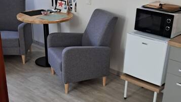 Espaço compacto para viver, com uma poltrona cinza, uma mesa redonda pequena com porta-retrato, micro-ondas e um armário branco.