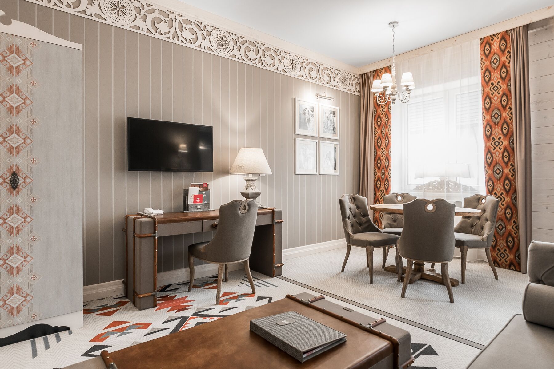 Quarto de hotel com televisão de tela plana, escrivaninha com luminária, área de refeições com cadeiras e cortinas estampadas.