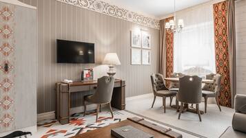 Quarto de hotel com televisão de tela plana, escrivaninha com luminária, área de refeições com cadeiras e cortinas estampadas.