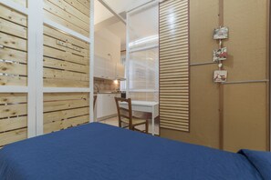 Quarto com cama, escrivaninha e cadeira. Há uma janela com persianas e uma prateleira fixada na parede com quadros.