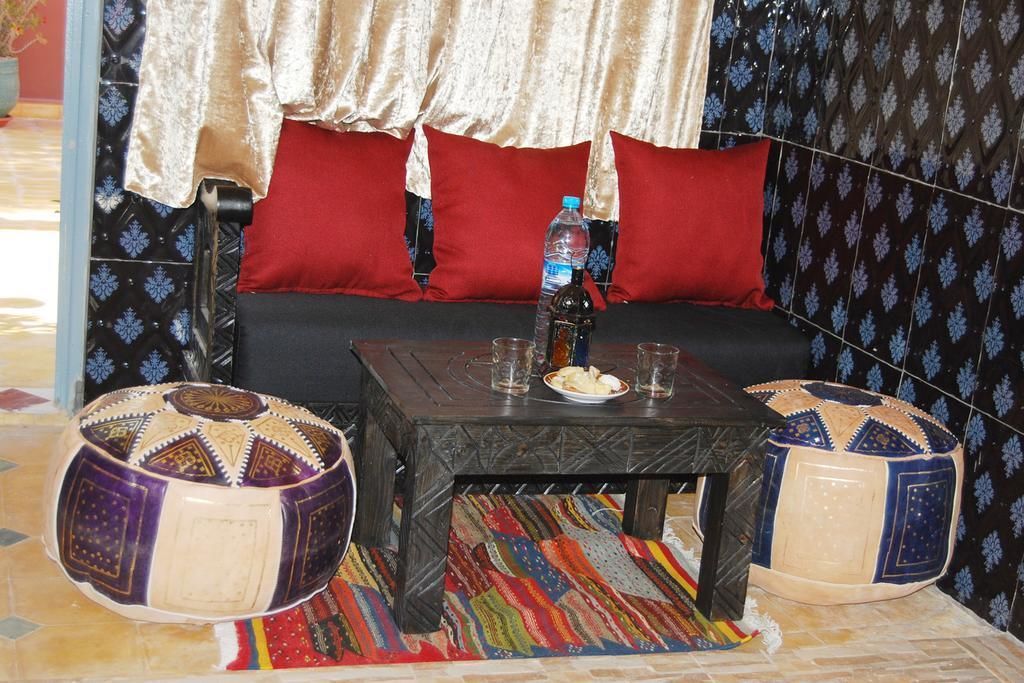 Um quarto aconchegante com um sofá preto, almofadas vermelhas, uma pequena mesa de madeira com uma garrafa e copos, e dois pufes decorativos.