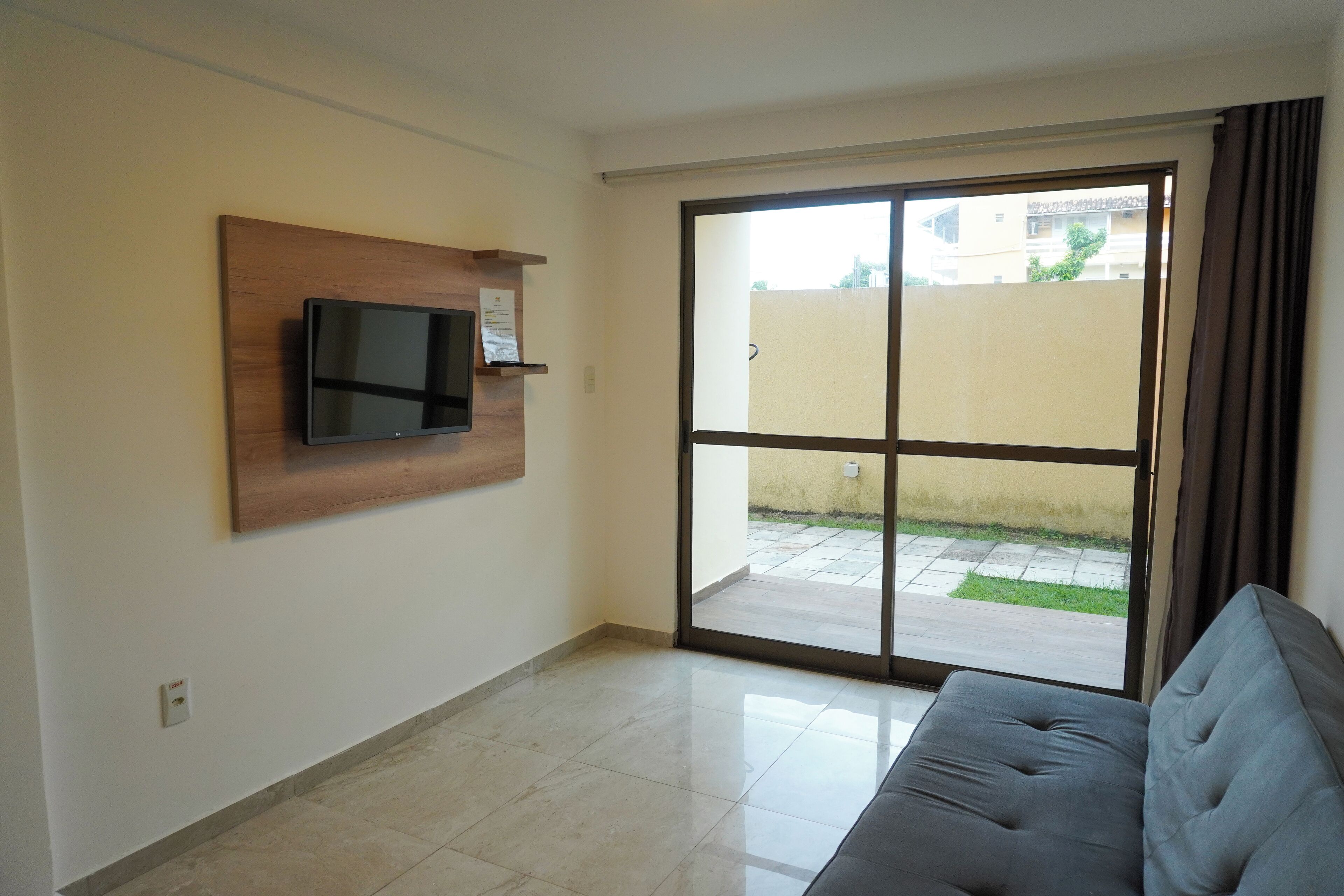 Sala de estar com televisão de tela plana presa a um painel de madeira, porta de vidro deslizante para área externa e sofá cinza.