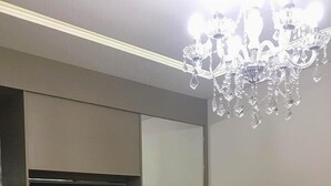 Uma sala de estar com televisão de tela plana sobre um suporte de madeira, uma geladeira pequena branca, um sofá com almofada estampada e um lustre.