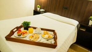Quarto de hotel com uma cama bem arrumada, uma bandeja com itens de café da manhã, incluindo frutas, doces e uma xícara de café.