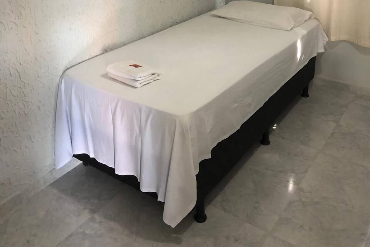 Quarto pequeno com uma cama de solteiro, edredom branco, cama bem arrumada com toalha dobrada e cortina na janela.