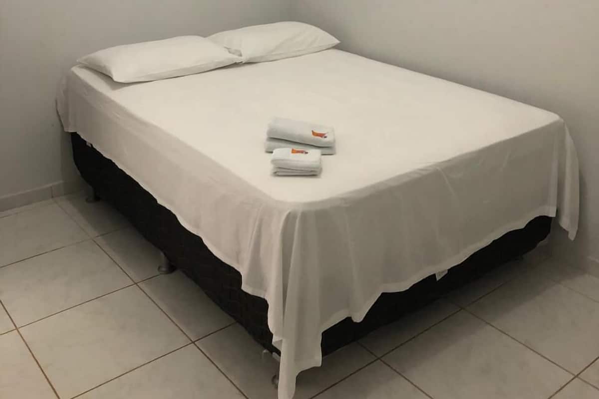 Uma cama pequena e bem arrumada, com lençóis brancos, em um quarto com paredes brancas e piso de azulejos.