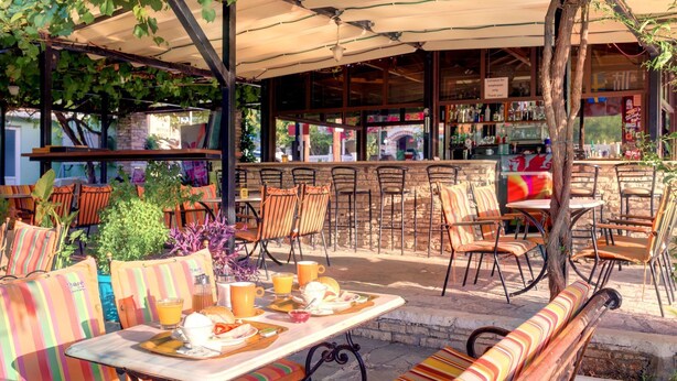 Um café ao ar livre com mesas, cadeiras, um espaço de bar e vista para a rua.