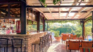 Um espaço de bar ao ar livre com paredes de pedra, banquetas de metal e cadeiras laranja.