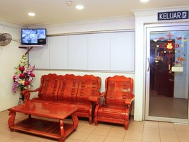 Uma área de recepção com um sofá de madeira, uma mesa pequena, uma televisão montada na parede e uma porta com uma placa que diz 'KELUAR'.