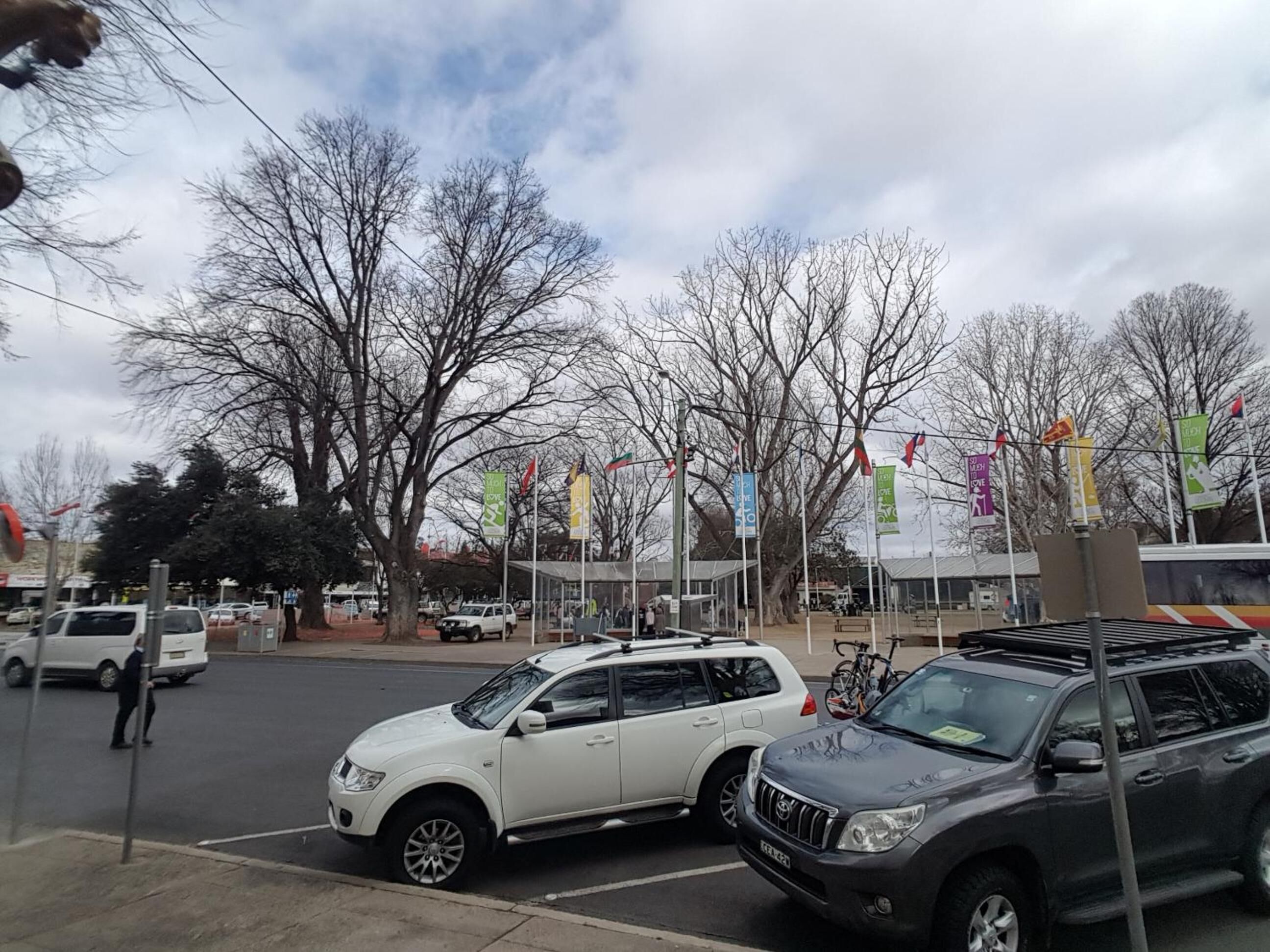Um estacionamento com vários carros, incluindo dois SUVs, um branco e um preto. Árvores e um edifício com bandeiras ao fundo.