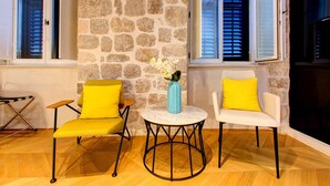 Quarto com parede de pedra, duas cadeiras com almofadas amarelas, uma mesa redonda pequena e um vaso com flores.