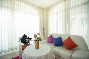 Uma sala de estar com sofá branco, mesa redonda com um vaso de flores e cortinas leves.