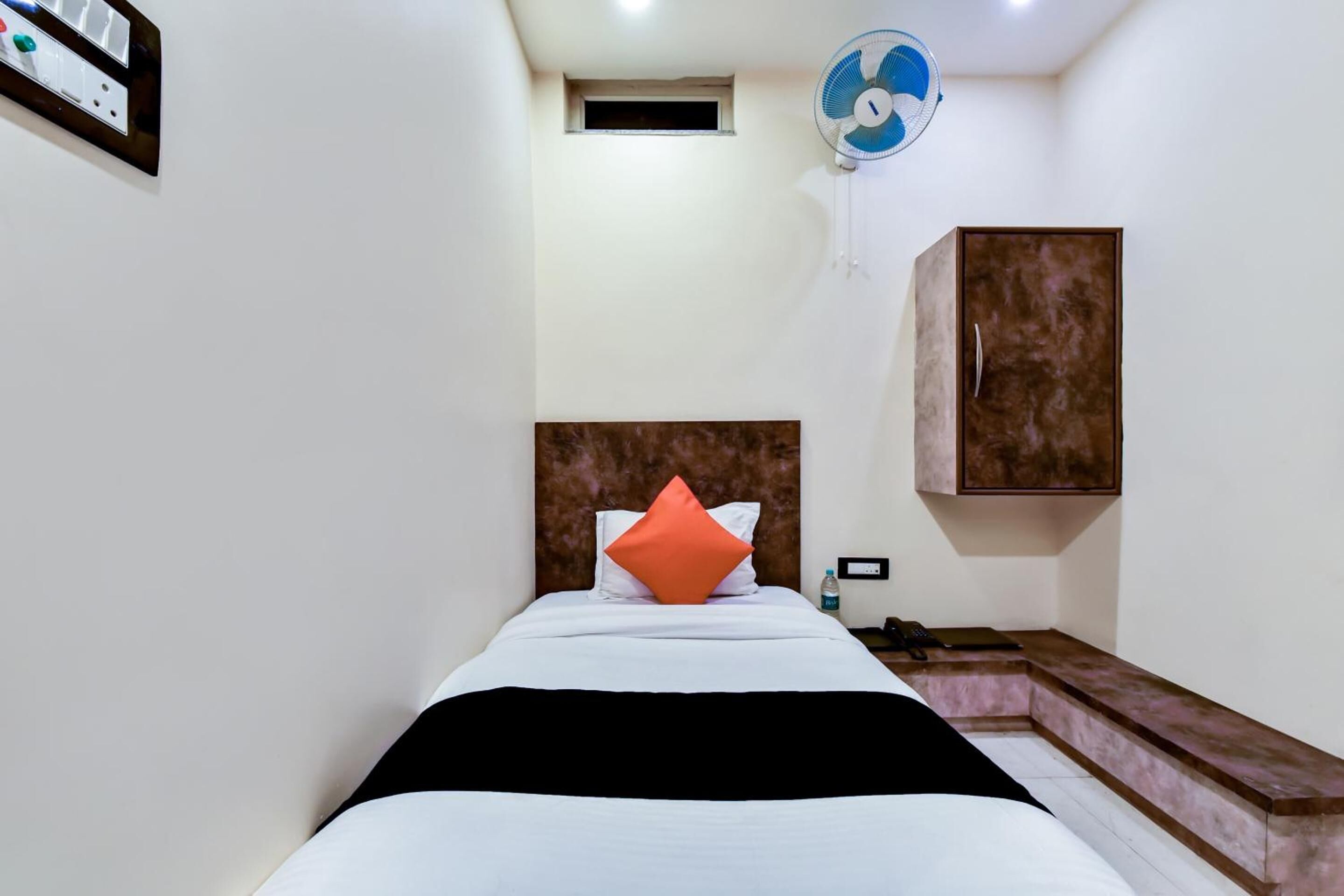 Quarto de hotel com cama, mesa de cabeceira, ventoinha na parede, uma pequena janela e um armário.