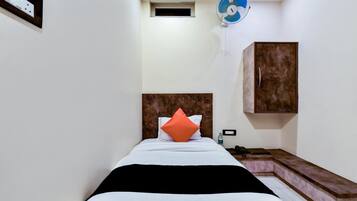 Quarto de hotel com cama, mesa de cabeceira, ventoinha na parede, uma pequena janela e um armário.
