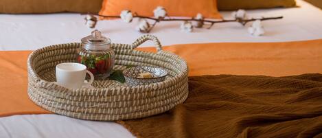 Cama com travesseiros laranja e castanhos, bandeja de palha com bule e xícara, e um mural na parede.