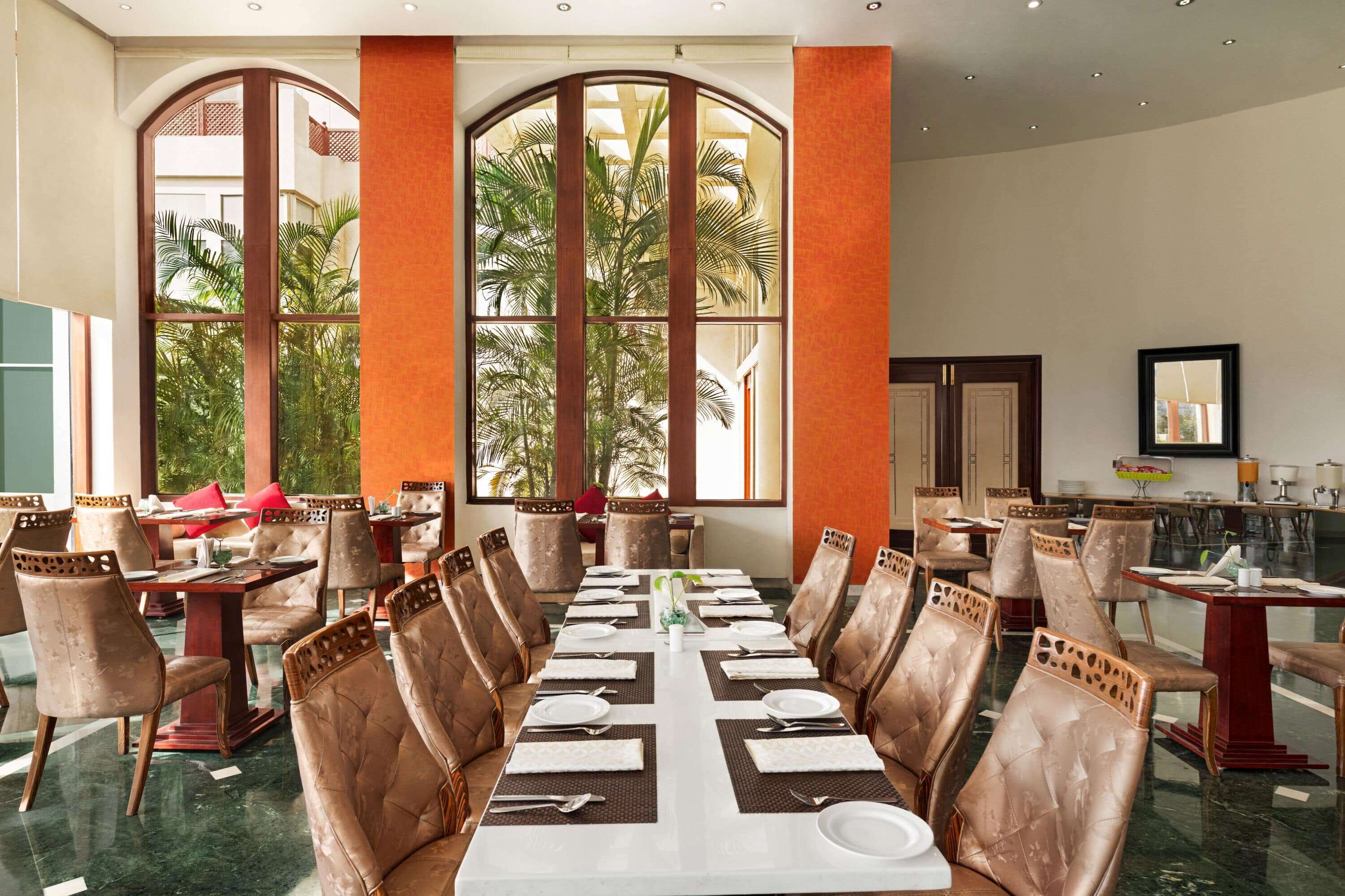 Uma ampla sala de jantar com cadeiras de encosto alto e uma mesa comprida posta para refeição, grandes janelas com vista para palmeiras, e design de interiores moderno.