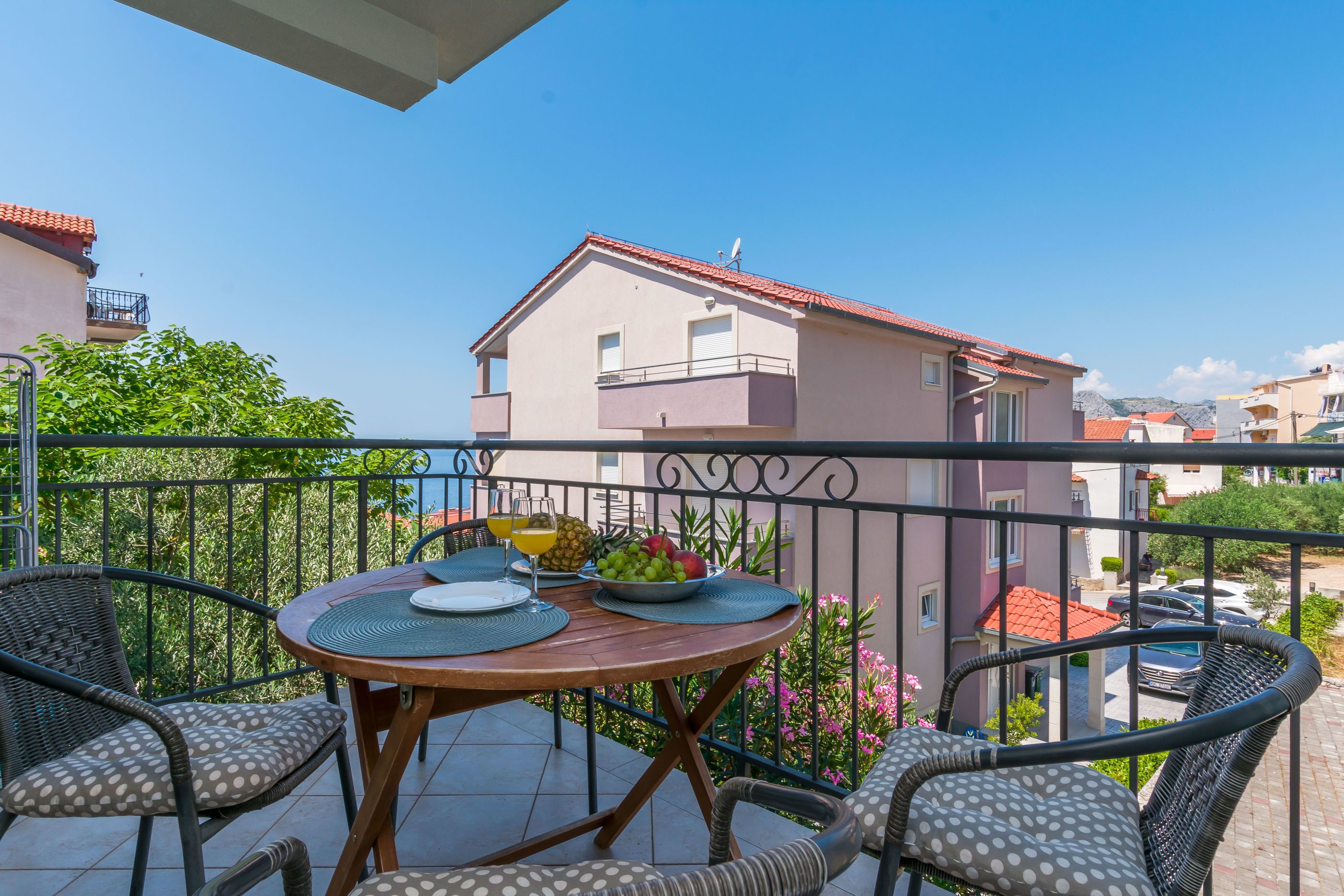 Varanda com mesa posta para dois, rodeada por cadeiras de vime, com vista para uma área residencial com casas e céu claro.