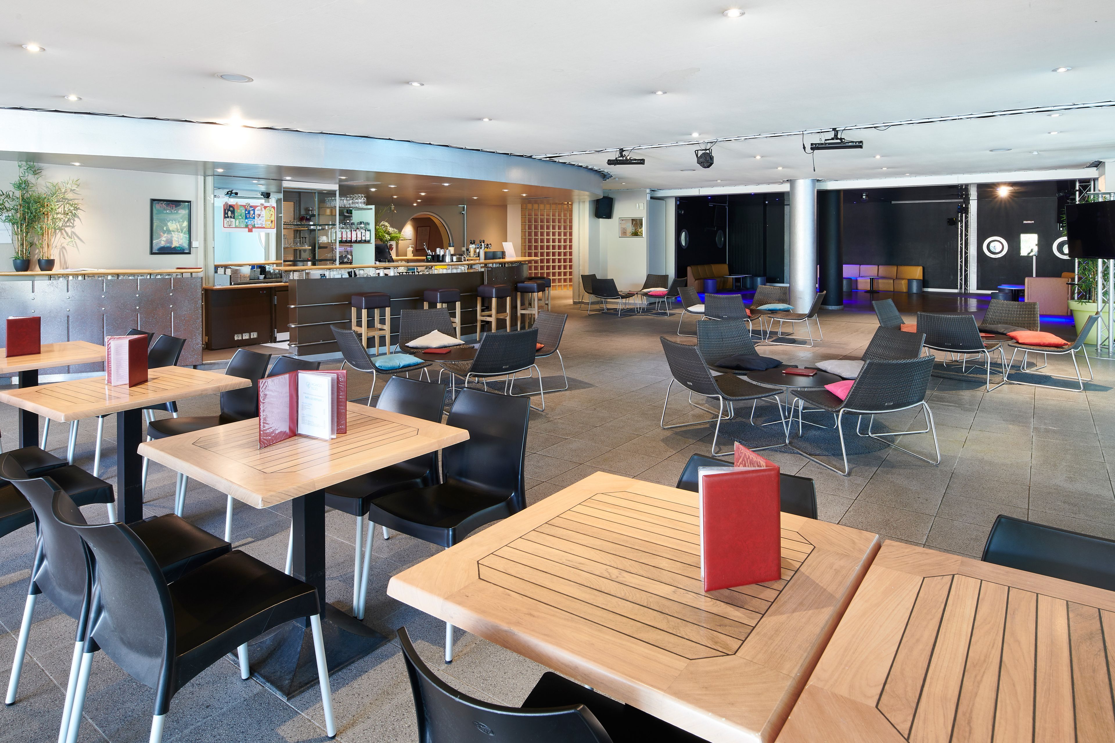 Um café moderno com mesas de madeira e cadeiras pretas, uma área de bar e um palco para apresentações.