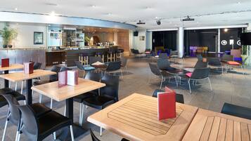 Um café moderno com mesas de madeira e cadeiras pretas, uma área de bar e um palco para apresentações.