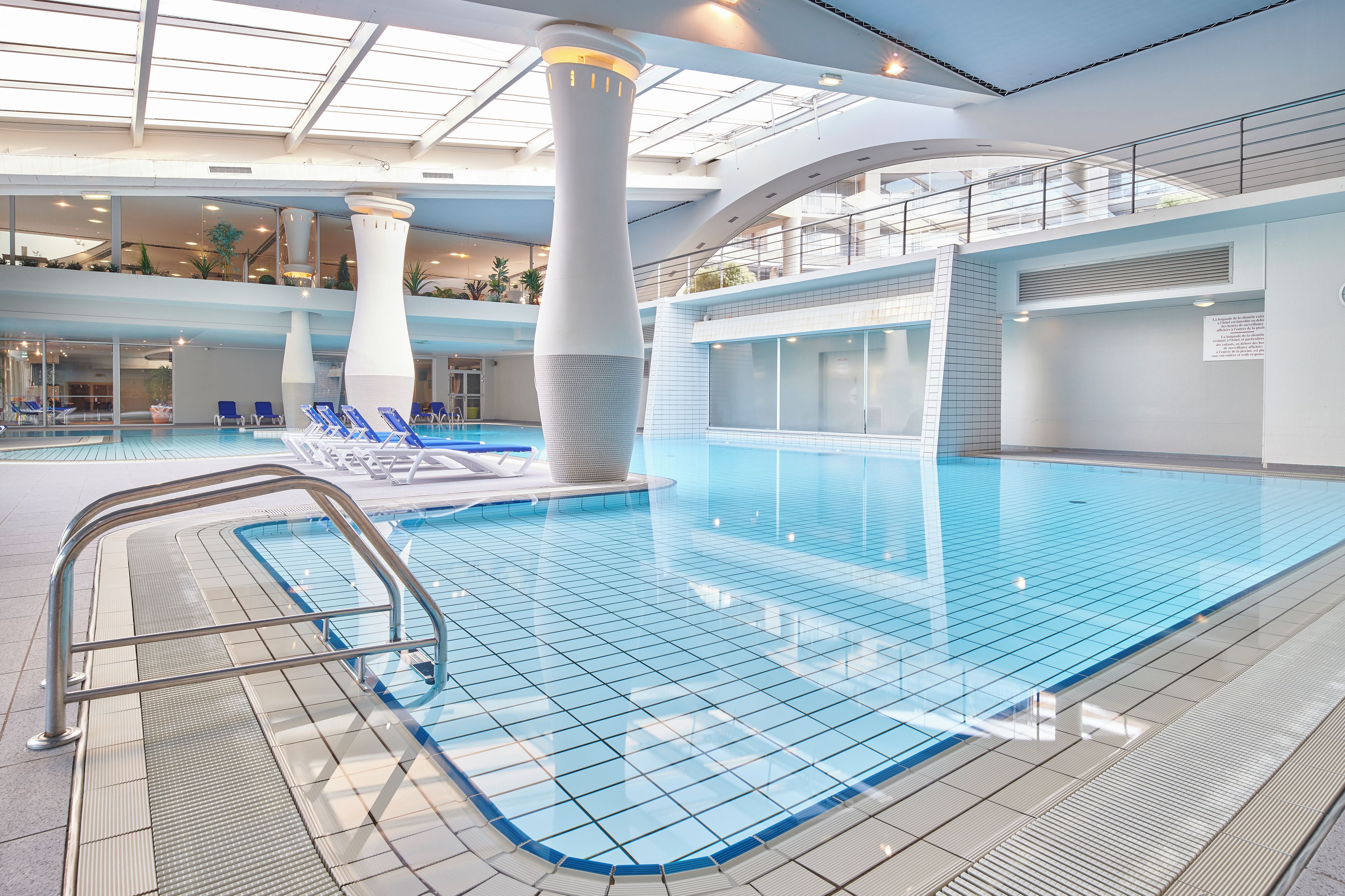 Piscina interior moderna com água azul cristalina, teto iluminado pelo sol e área de lazer com espreguiçadeiras.