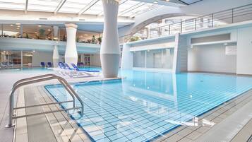 Piscina interior moderna com água azul cristalina, teto iluminado pelo sol e área de lazer com espreguiçadeiras.