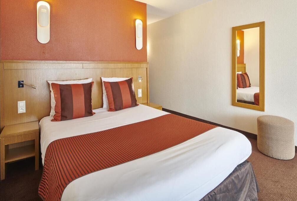 Quarto de hotel com uma cama grande, duas mesas de cabeceira, um espelho e um pequeno banquinho.