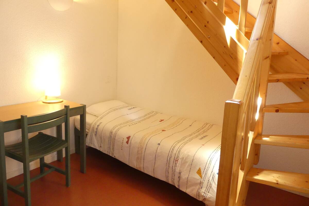 Quarto pequeno com cama de solteiro, escada de madeira, escrivaninha com cadeira e luminária na parede.