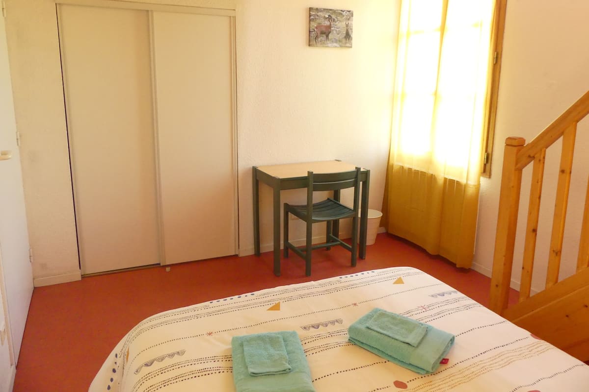 Quarto com cama, escrivaninha e cadeira. Há uma porta e uma janela com cortinas.