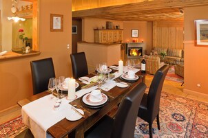 Uma sala de jantar com uma longa mesa de madeira posta para refeição, uma lareira e uma área de estar aconchegante com um tapete estampado.