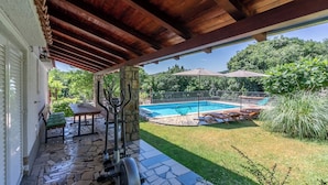 Um alpendre coberto com piso de pedra, equipamentos de exercícios ao ar livre, piscina e vista para um jardim exuberante.