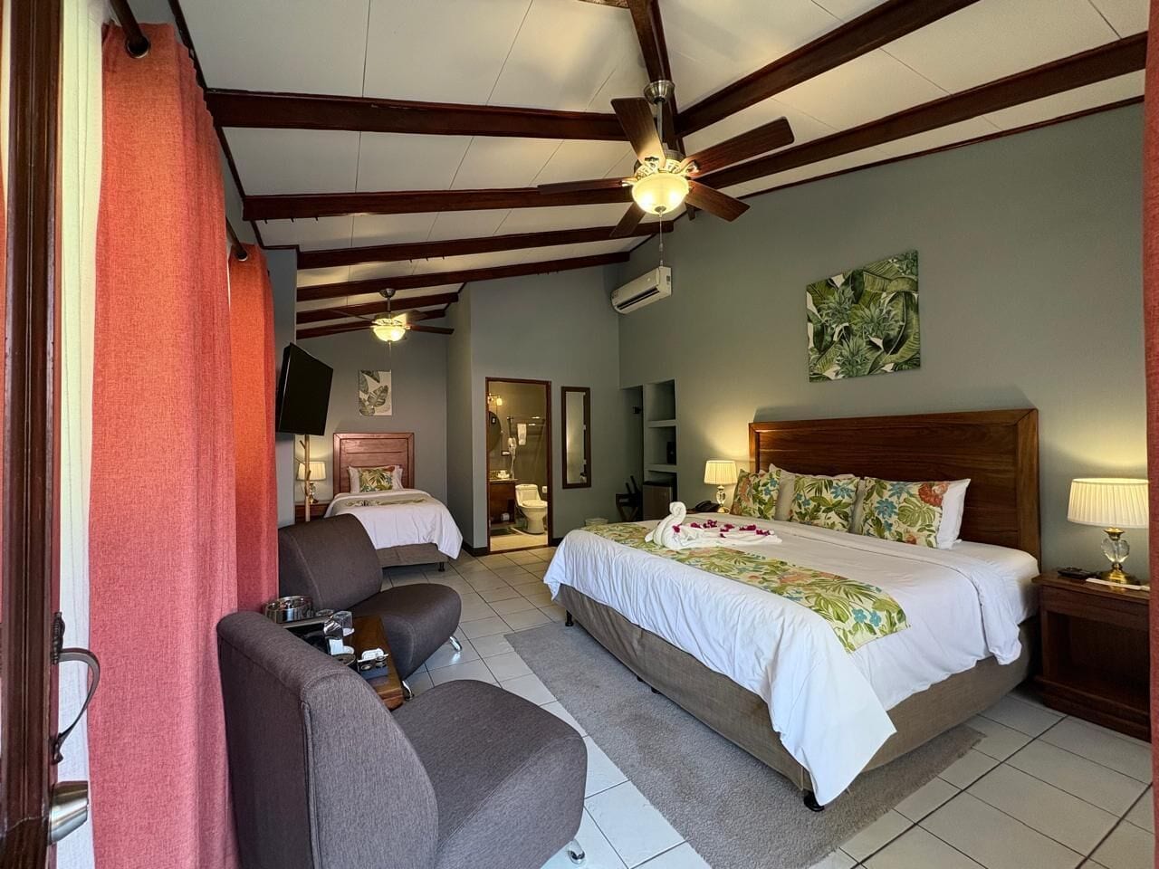 Quarto de hotel com uma cama grande, uma escrivaninha, uma cadeira, um ventilador de teto e um banheiro visível por uma porta aberta.