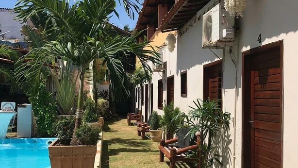 Uma área de piscina com palmeira e plantas em vasos, um deck de madeira e um edifício com telhado telhado e portas de madeira.