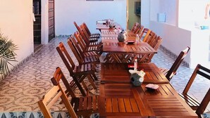 Uma área de refeições ao ar livre coberta, com mesas e cadeiras de madeira, uma grande flor decorativa e piso de azulejos.