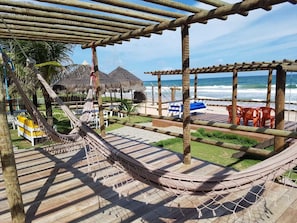 Uma rede sob uma pérgola de madeira com vista para uma praia com guarda-sóis de palha e uma área de piscina.