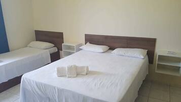 Quarto de hotel com duas camas de solteiro e uma cama de casal, todas com roupa de cama branca. Há duas mesas de cabeceira, uma em cada cama de solteiro e outra na cama de casal.