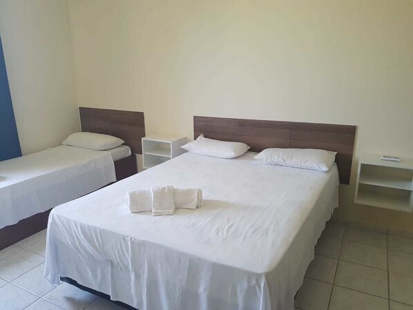 Quarto de hotel com duas camas de solteiro e uma cama de casal, todas com roupa de cama branca. Há duas mesas de cabeceira, uma em cada cama de solteiro e outra na cama de casal.