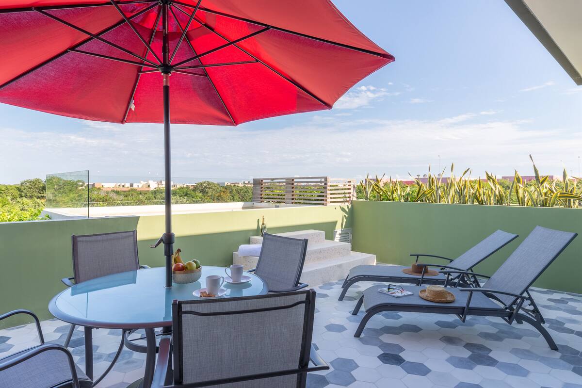 Um pátio com mesa de vidro, cadeiras, um guarda-sol vermelho e vista para a cidade.