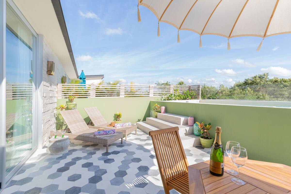 Uma varanda com chão de azulejos hexagonais, mobiliário de vime, mesa de madeira com garrafa e copos, e vista para vegetação e um edifício ao fundo.