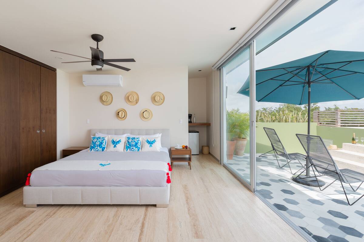 Quarto moderno com cama grande, ventilador de teto e vista para um pátio externo com cadeiras e mesa.