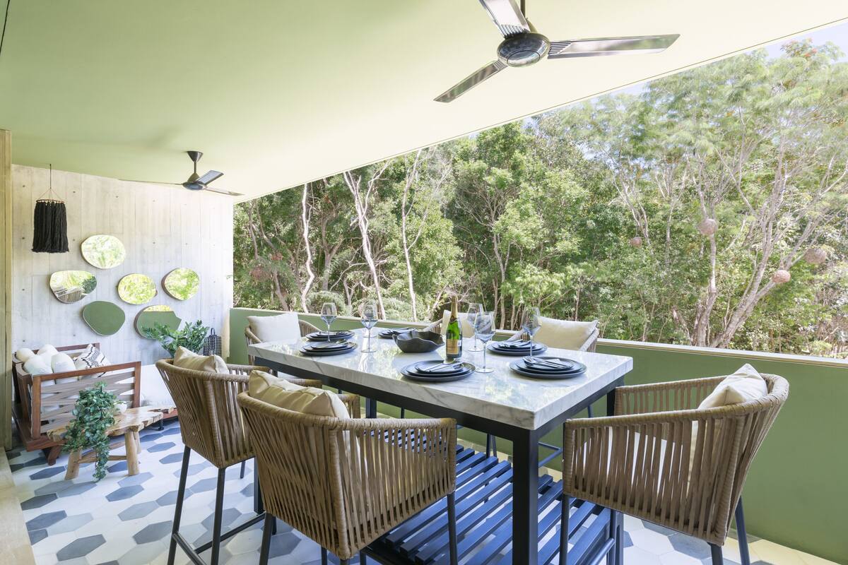 Uma área de refeições com mesa posta para seis pessoas, rodeada por cadeiras de vime, um ventilador de teto e vista para árvores.